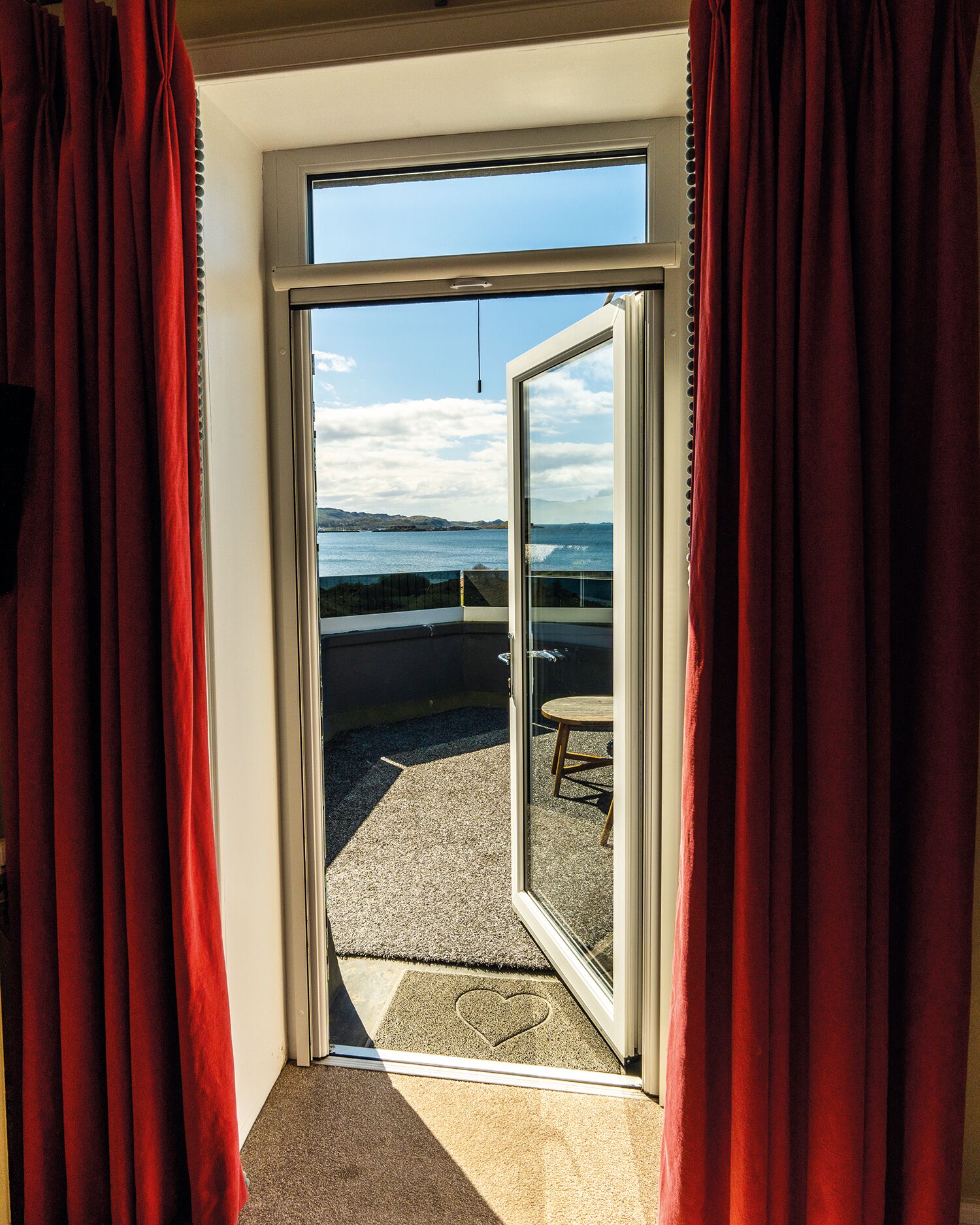 Uma porta de vidro deslizante dá para uma varanda com vista para o mar e colinas ao longe.