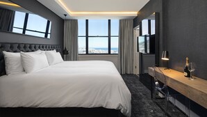 Quarto de hotel moderno com uma cama grande, uma escrivaninha de madeira, uma televisão e vista para a cidade.