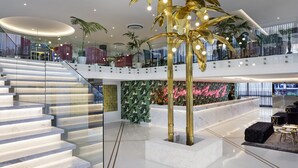 Um hall de hotel moderno com uma escadaria central, um balcão de recepção e uma grande planta decorativa.