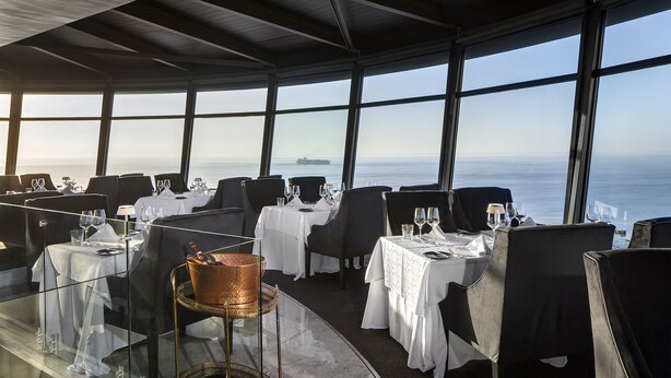 Restaurante com mesas preparadas para refeições, amplas janelas com vista para o mar e guarda-corpo de vidro separando o salão do visual.