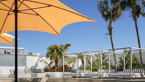 Uma área à beira da piscina com espreguiçadeiras, uma mesa com garrafa e copo, e um grande guarda-sol.
