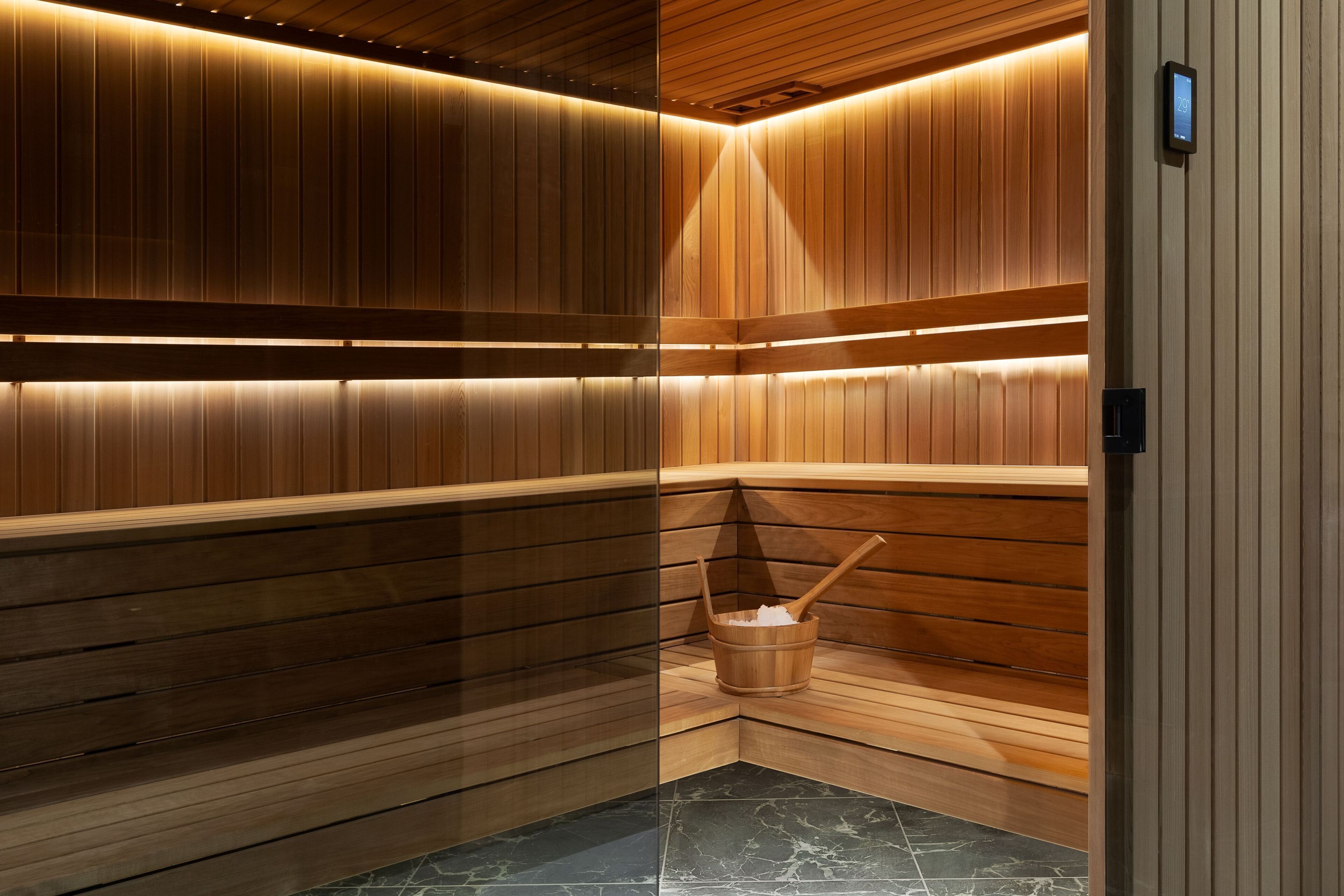 Uma sauna com bancos de madeira, um balde com escova e um painel digital na parede.