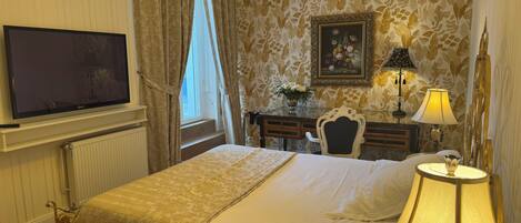 Quarto de hotel com cama, televisão, secretária, cadeira, abajur e janela com cortinas.