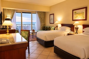 Quarto de hotel com duas camas, uma mesa de trabalho com telefone, uma cadeira, uma mesinha com frutas e uma varanda com vista.