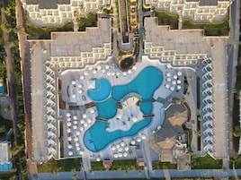 Vista aérea de um hotel com uma grande piscina central, várias piscinas e uma área de jardim bem cuidada.