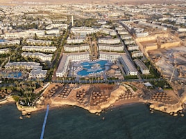 Vista aérea de um resort com vários edifícios, uma área de piscina central e uma praia com espreguiçadeiras.