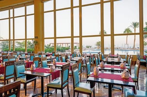 Um restaurante com várias mesas e cadeiras, janelas amplas e vista para uma piscina e palmeiras.