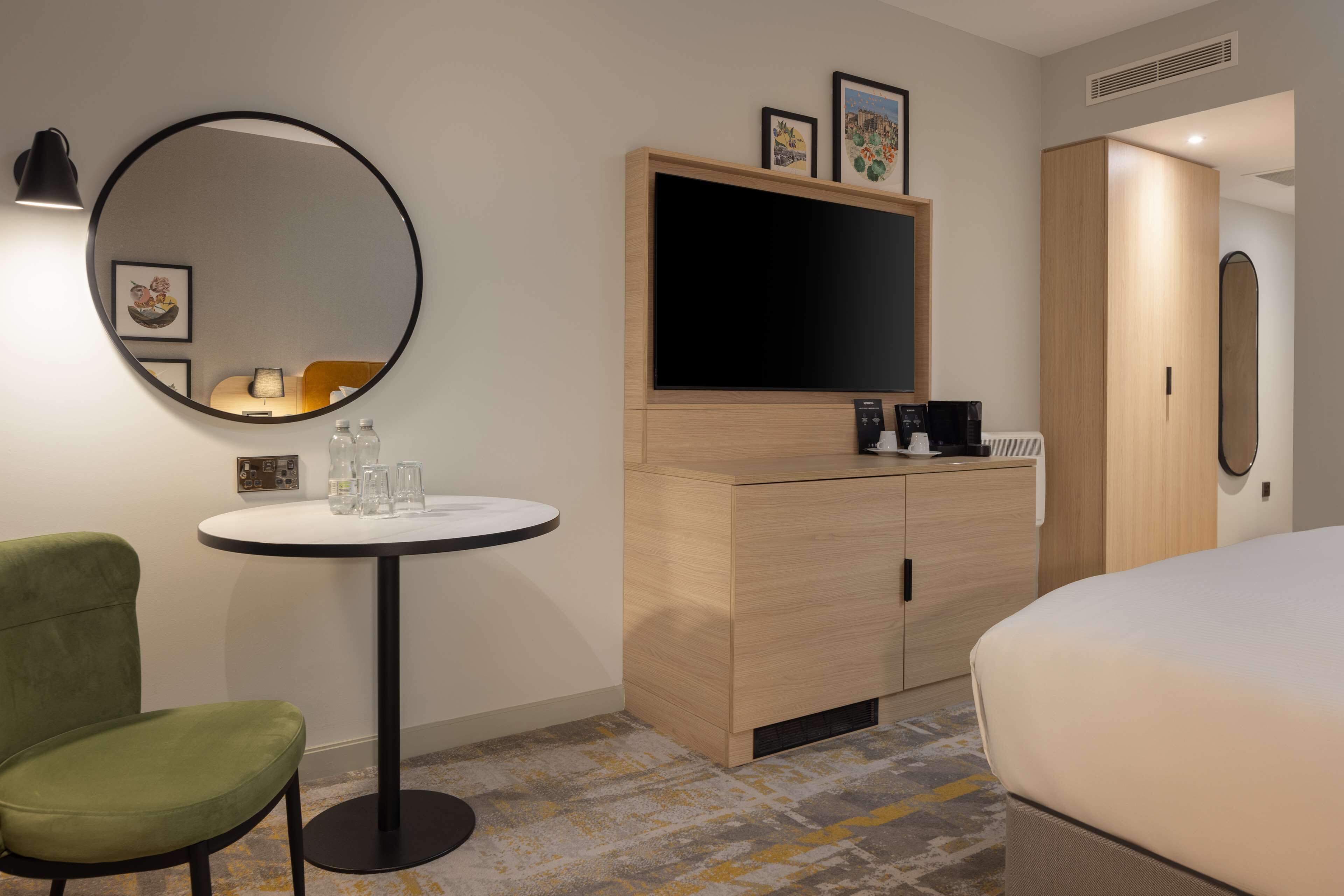 Quarto de hotel com cama, espelho redondo, televisão sobre um móvel de madeira, mesa pequena redonda com uma cadeira e luminária fixada na parede.