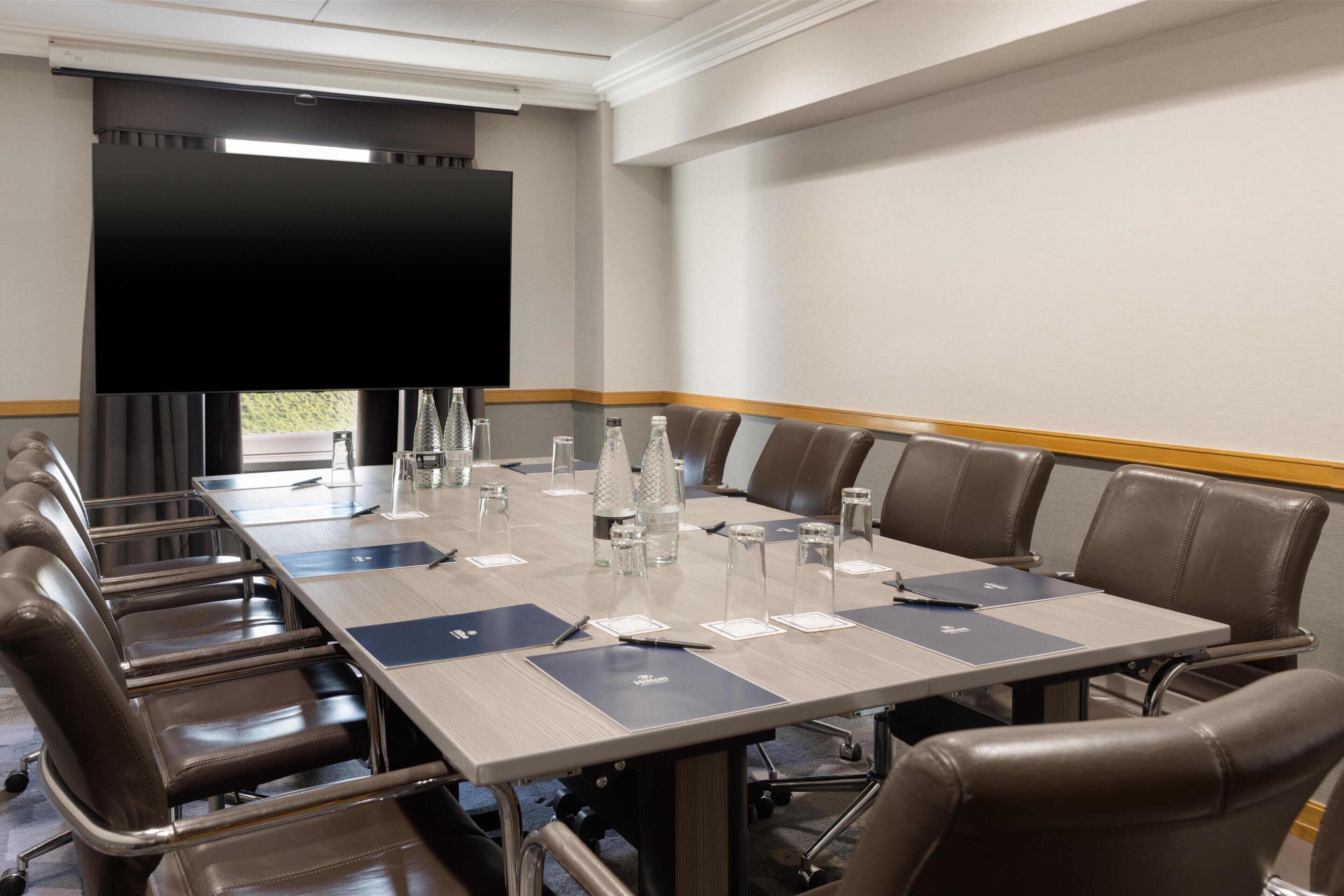 Sala de conferência com uma mesa comprida, cadeiras de couro marrom, garrafas de água e uma tela grande.