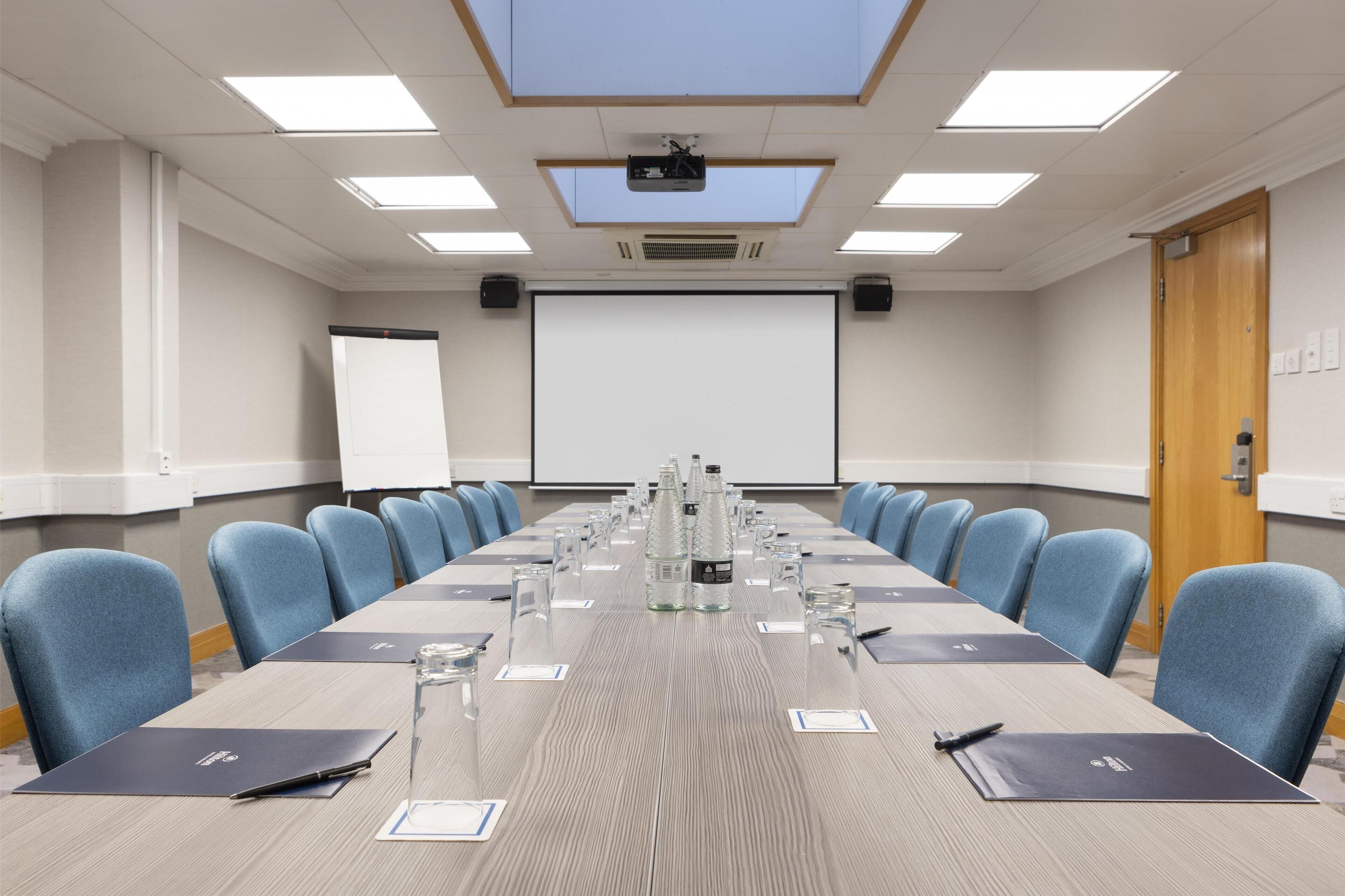 Uma sala de conferências com uma mesa comprida, cadeiras azuis, um projetor e uma lousa.