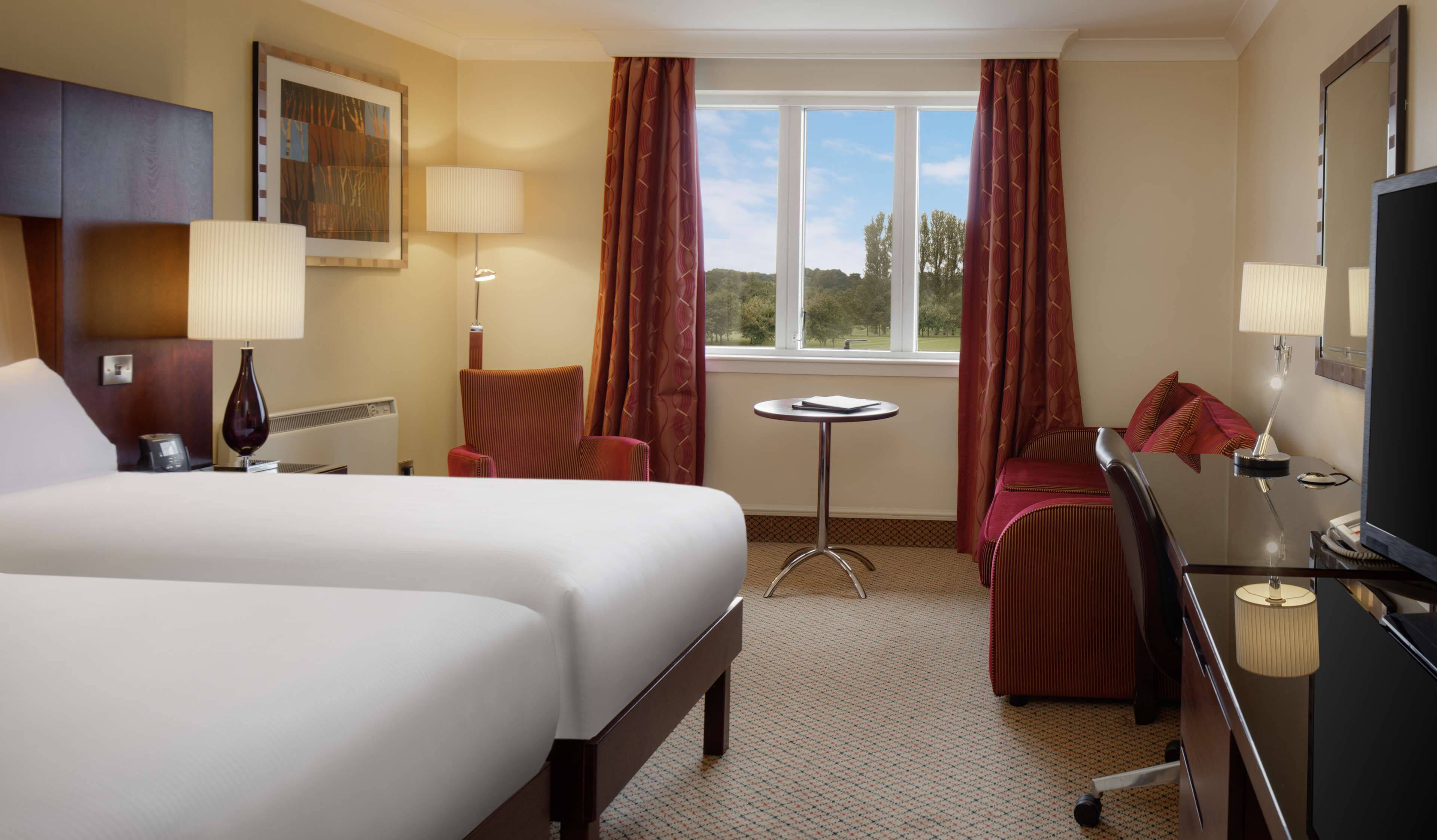 Quarto de hotel com duas camas, uma escrivaninha, uma cadeira, uma mesinha e uma janela com cortinas.