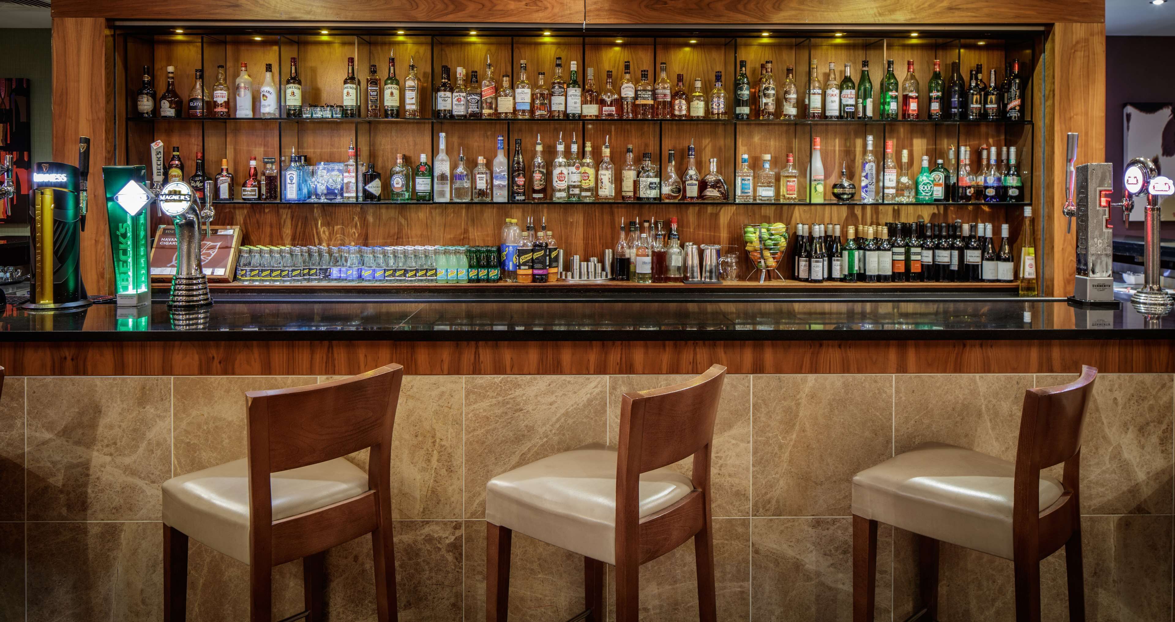 Um bar com várias prateleiras cheias de garrafas de bebidas, um balcão e três banquetas de madeira.