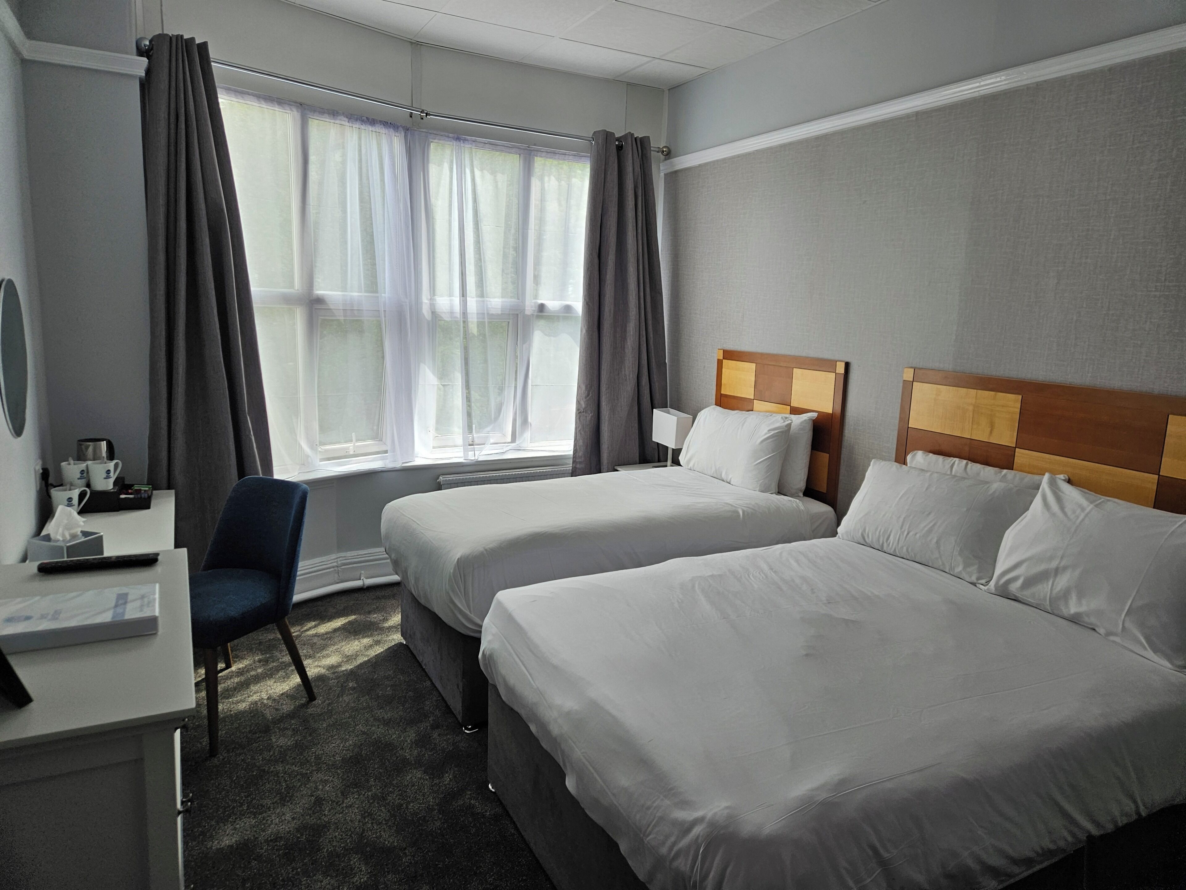 Quarto de hotel com duas camas, uma escrivaninha, uma cadeira, janela com cortinas e abajur.