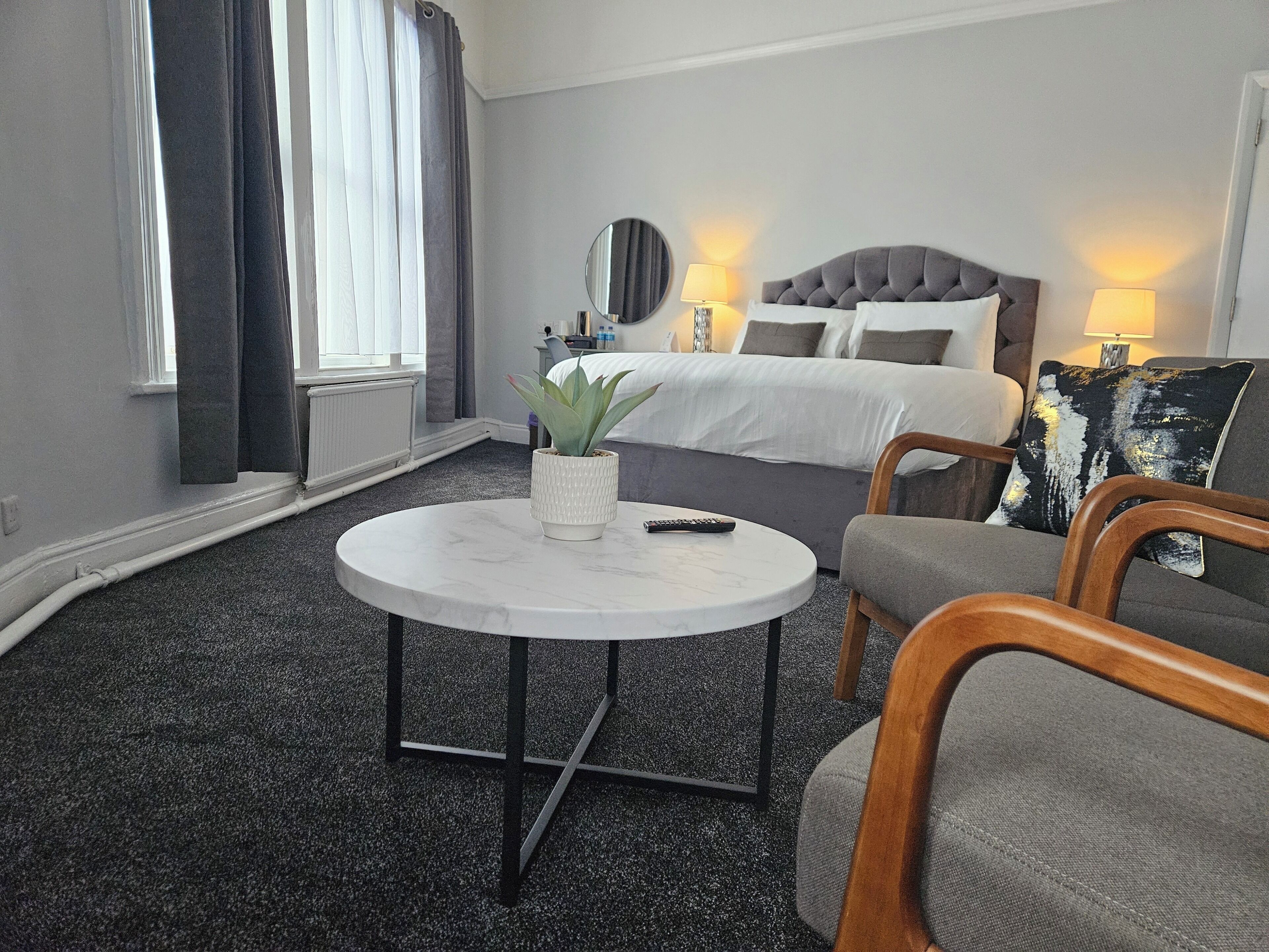 Quarto de hotel com uma cama, uma mesa redonda com uma planta, duas cadeiras, um abajur e uma janela com cortinas.