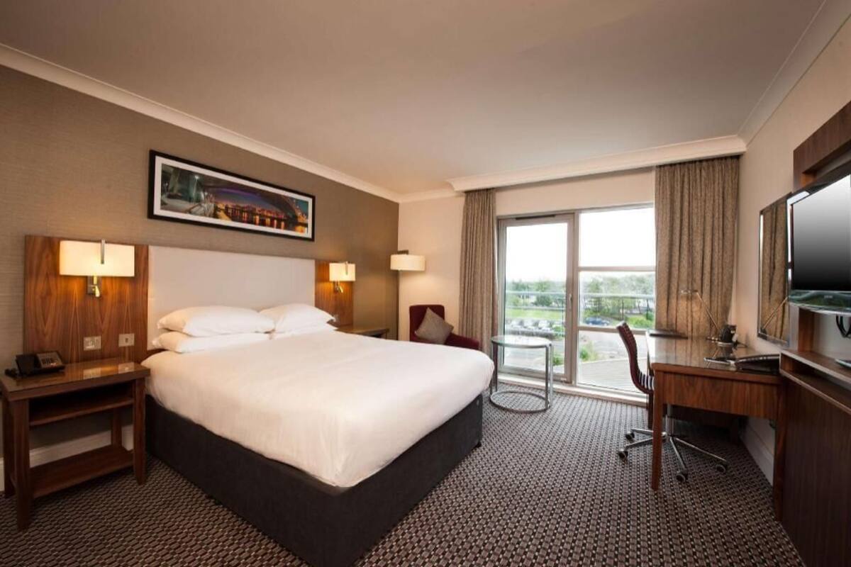 Quarto de hotel com uma cama grande, duas mesas de cabeceira com luminárias, uma escrivaninha com cadeira, televisão e uma janela ampla com cortinas.