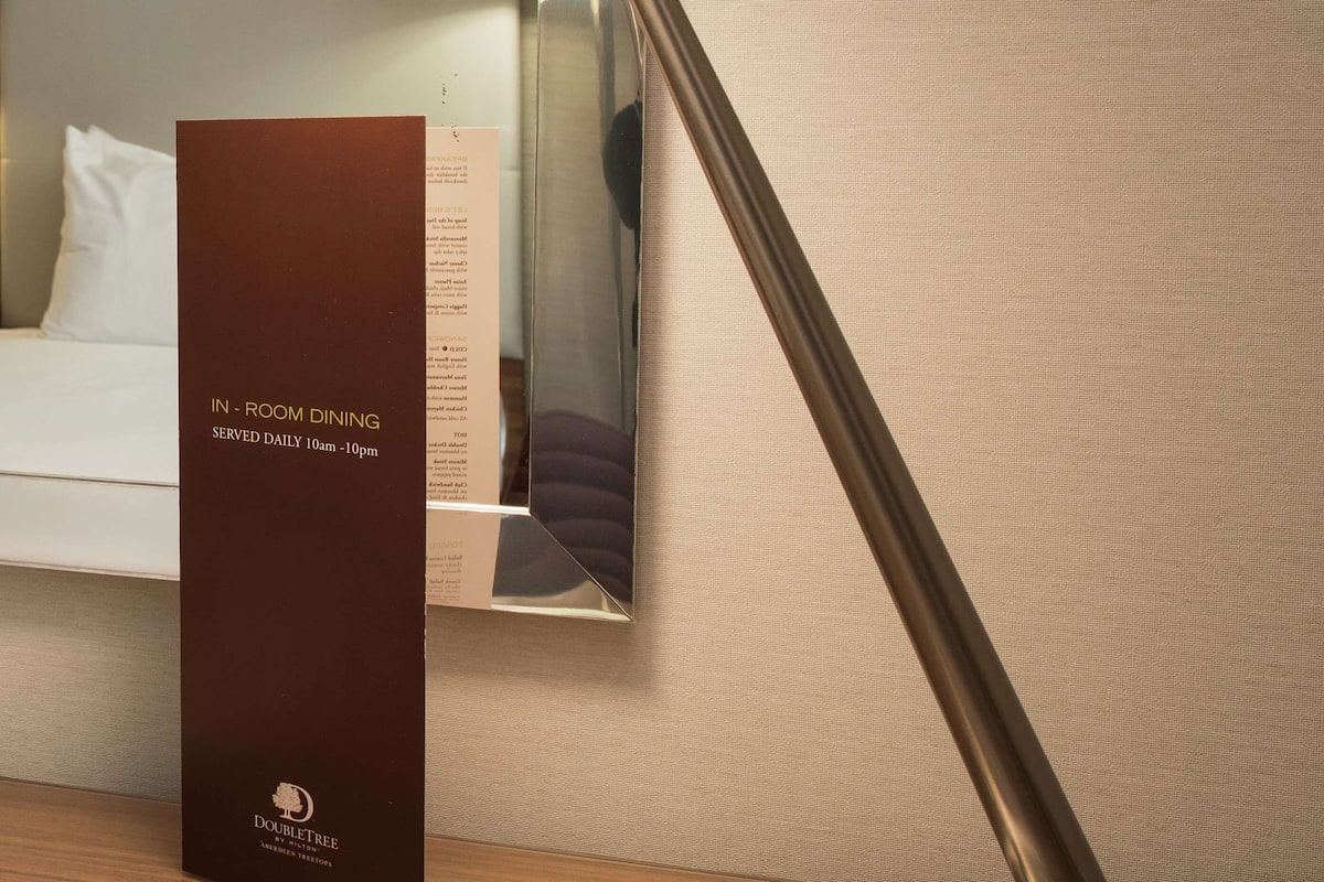 Quarto de hotel moderno com abajur, menu emoldurado e logotipo DoubleTree em um cartão marrom.