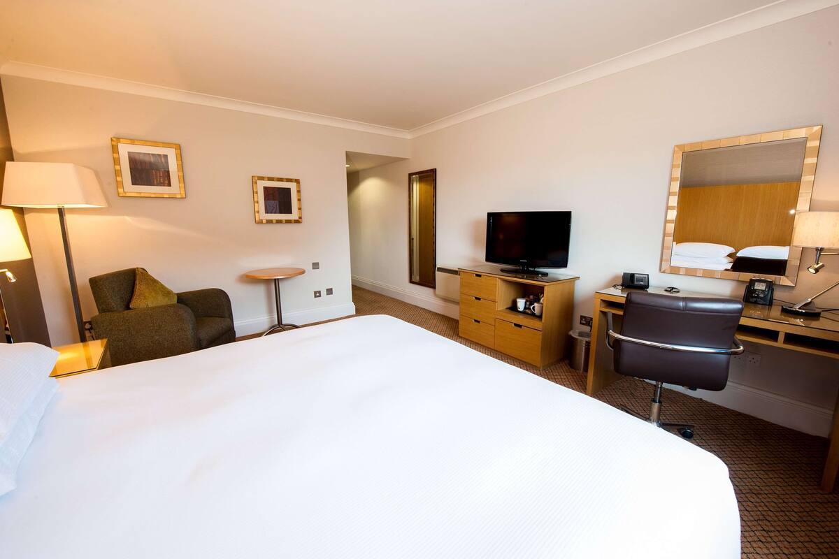 Quarto de hotel com uma cama grande, uma escrivaninha com computador, televisão, cadeira, abajur e cômoda.