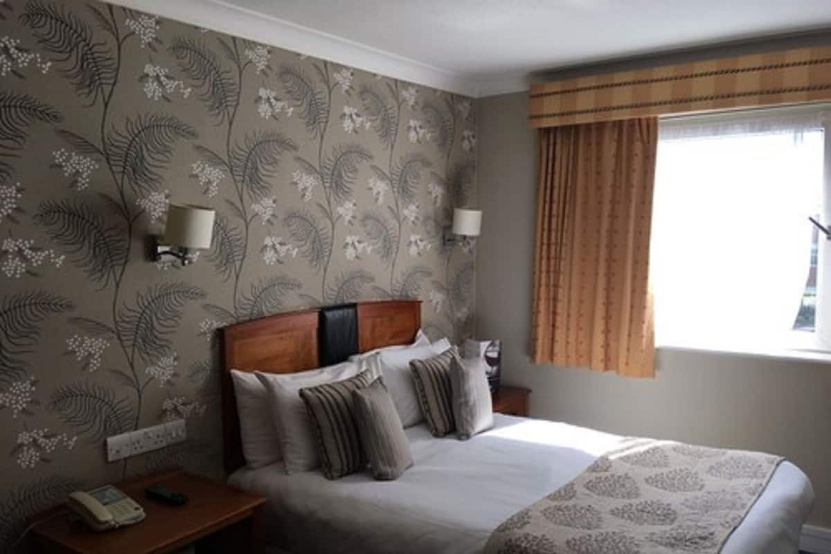 Quarto de hotel com cama, duas mesas de cabeceira, criado-mudo, telefone e janela com cortinas.