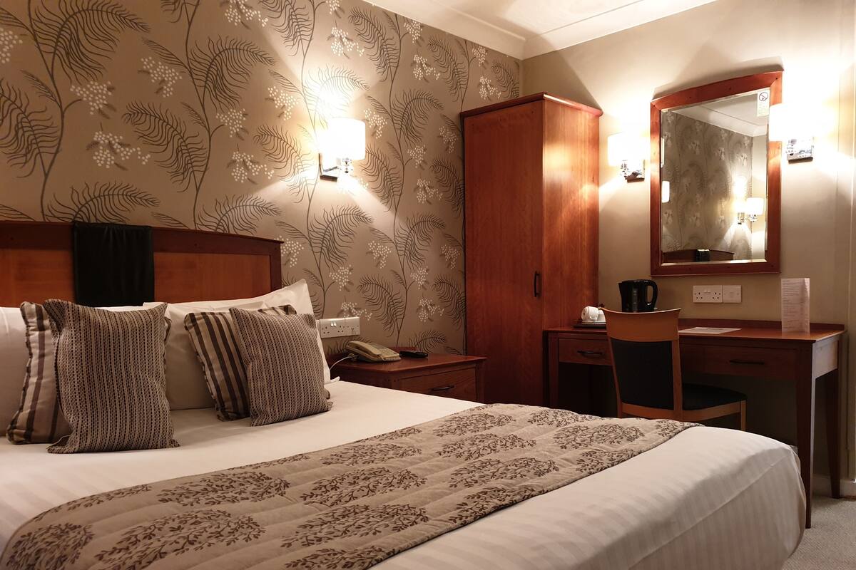 Quarto de hotel com cama, escrivaninha, cadeira, guarda-roupa e espelho.