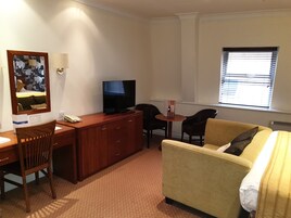 Quarto de hotel com secretária de madeira, televisão, sofá e pequena mesa com cadeiras.