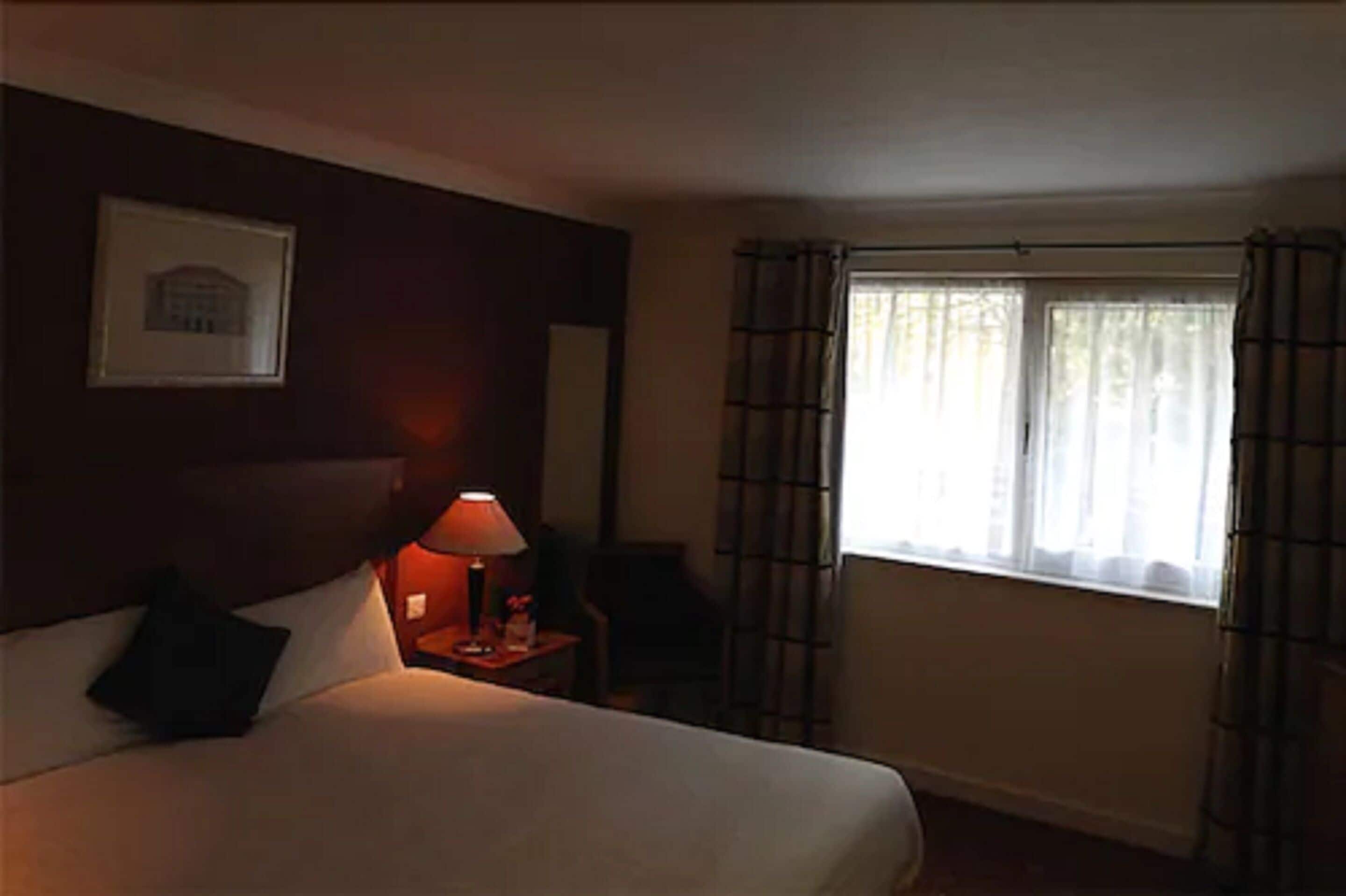 Quarto de hotel com cama, mesa de cabeceira com luminária, cadeira e janela com cortinas.