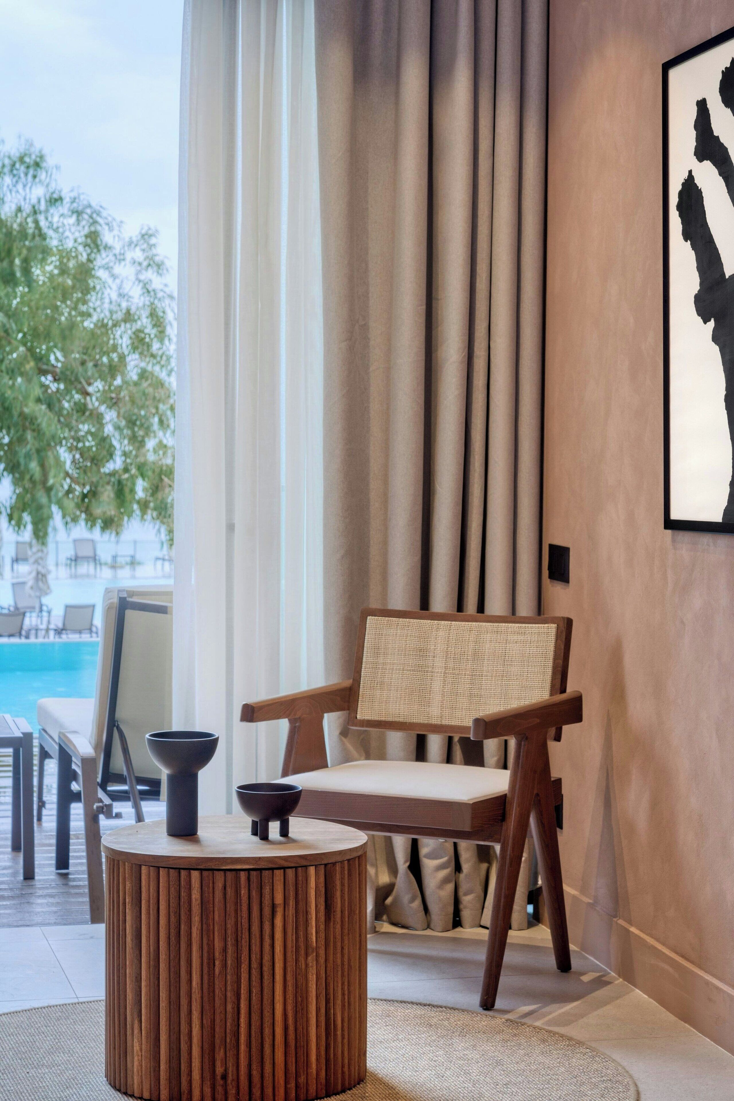 Quarto moderno com uma mesa redonda de madeira, uma cadeira de vime e vista para uma piscina e vegetação lá fora.