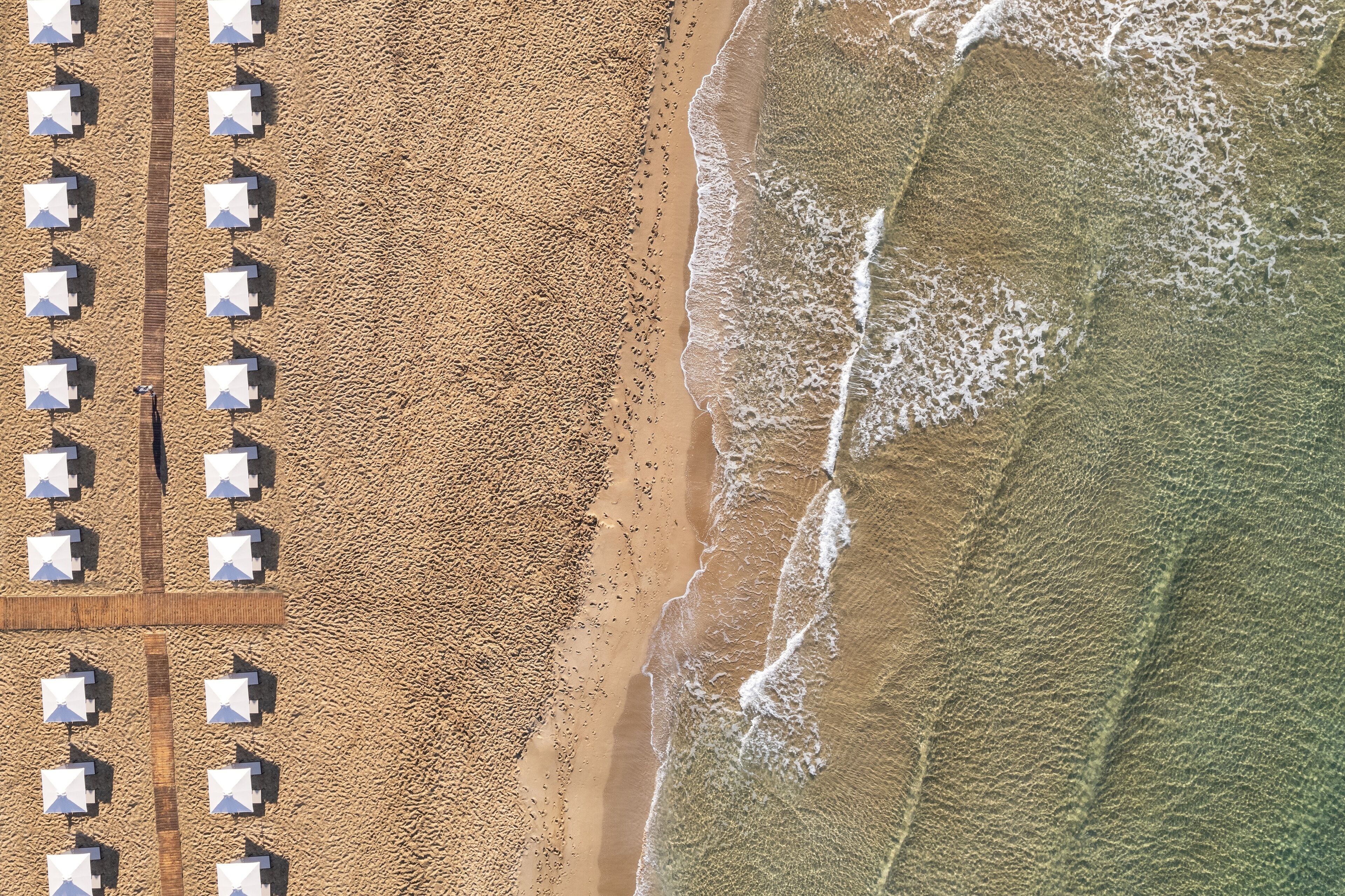 Vista aérea de uma praia com fileiras de guarda-sóis brancos e um calçadão de madeira.