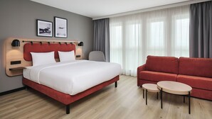 Quarto de hotel com uma cama, um sofá vermelho, duas mesinhas e uma janela com cortinas.