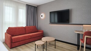 Quarto de hotel moderno com um sofá vermelho, duas mesinhas redondas e uma televisão fixada na parede, além de uma janela grande com cortinas leves.