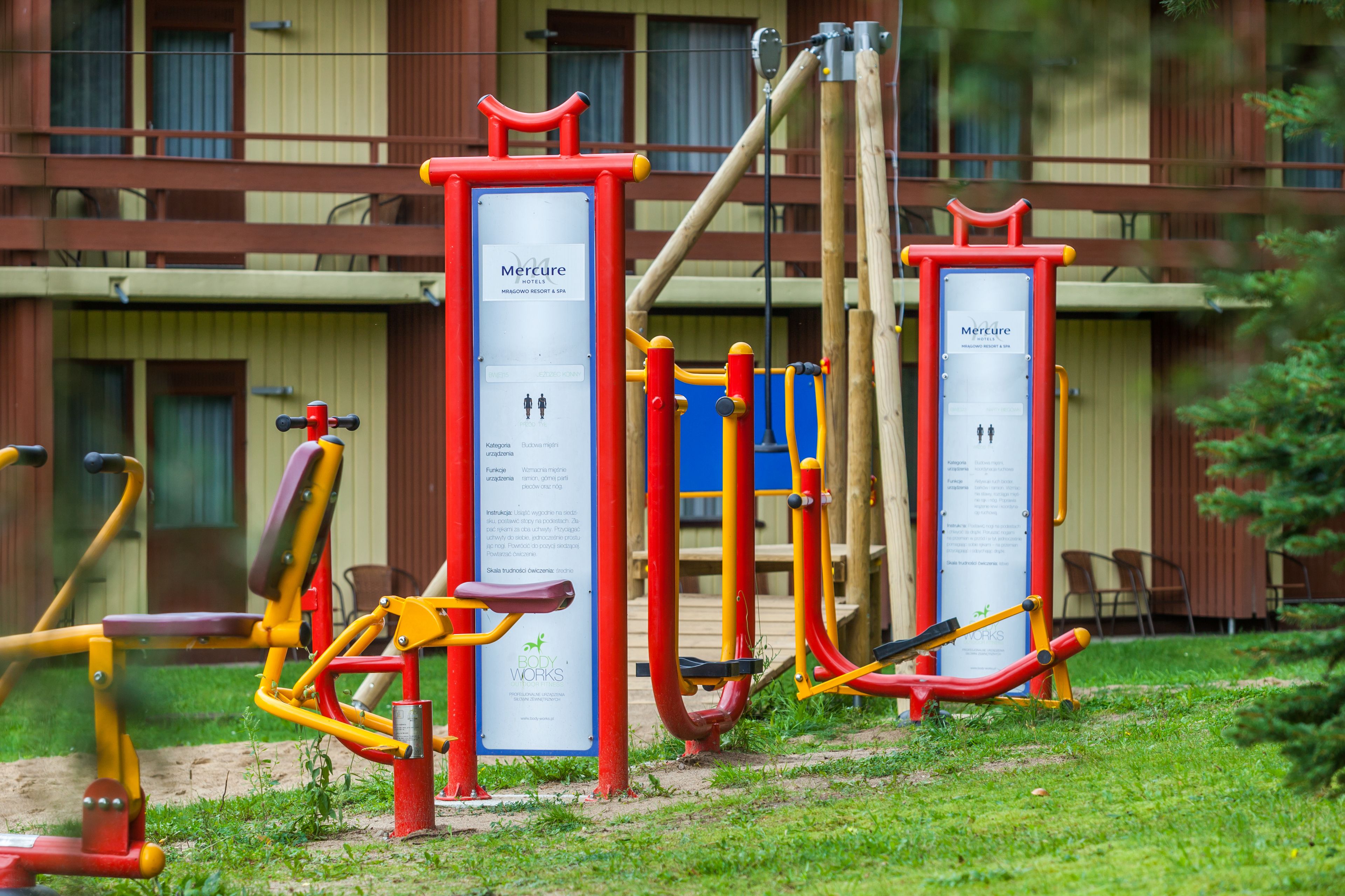 Equipamentos de ginástica ao ar livre com placas informativas, em frente a um edifício com varandas.