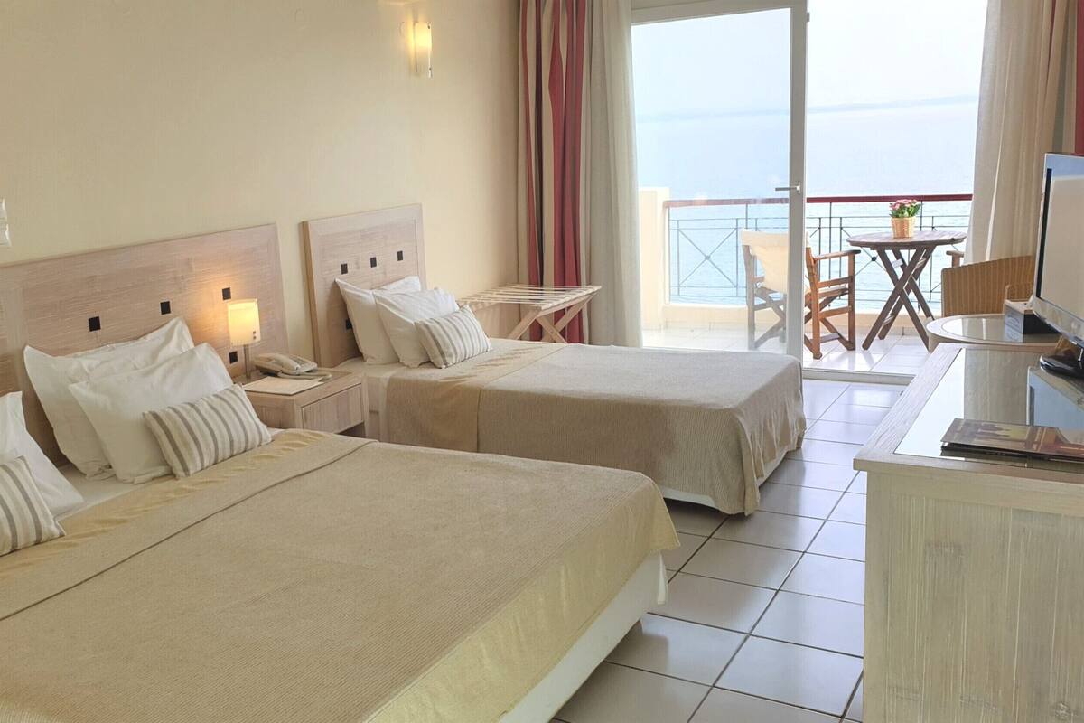 Quarto de hotel com duas camas, uma mesinha com cadeiras, televisão e varanda com vista para o mar.