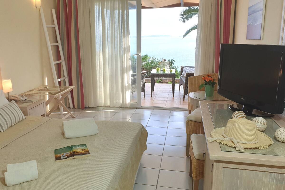 Quarto de hotel com cama, televisão, escrivaninha e vista para o mar.