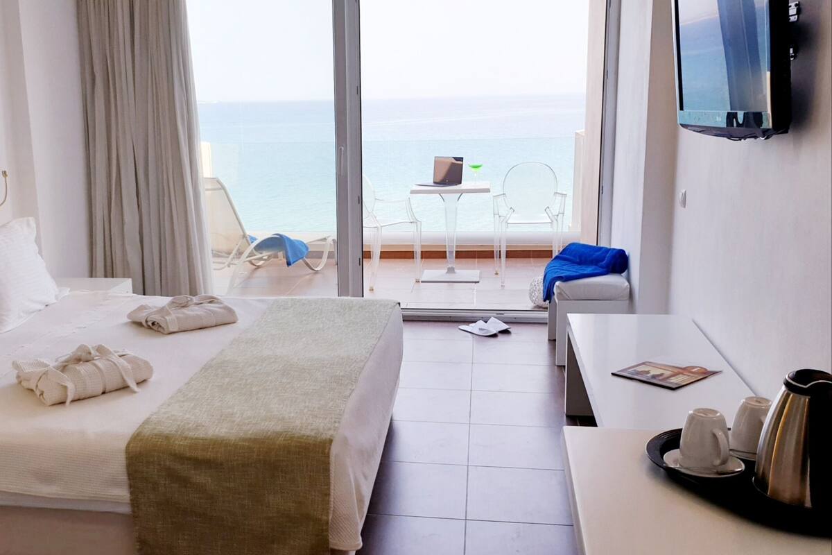 Quarto de hotel com uma cama, uma mesa com uma bandeja de copos, uma televisão e vista para o mar.