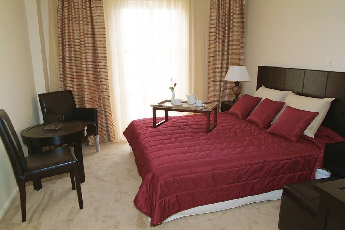 Quarto de hotel com uma cama grande, uma cadeira, uma mesa pequena com abajur e cortinas.