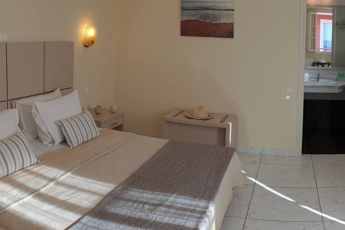 Quarto de hotel com uma cama, mesas de cabeceira, televisão, banheiro visível por uma porta aberta e um quadro na parede.