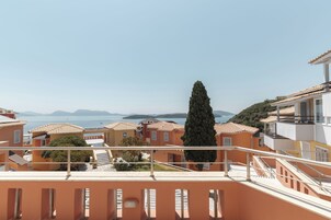 Um bairro residencial com casas de telhado em terracota, uma cipreste imponente e vista para o mar e montanhas ao longe.