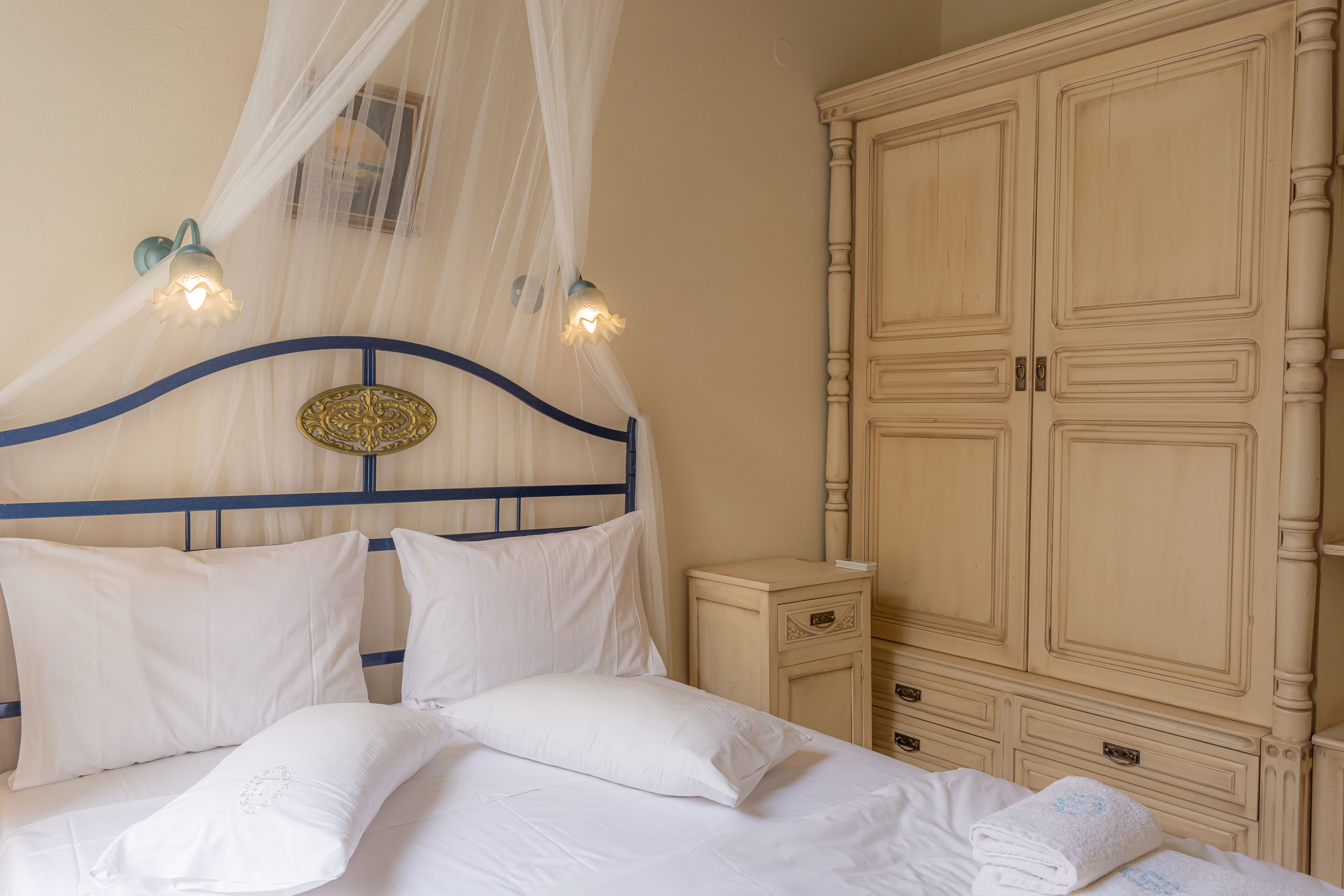 Cama com lençóis brancos, dossel e luminárias laterais. Há um guarda-roupa de madeira com várias portas e uma pequena mesa de cabeceira com abajur.
