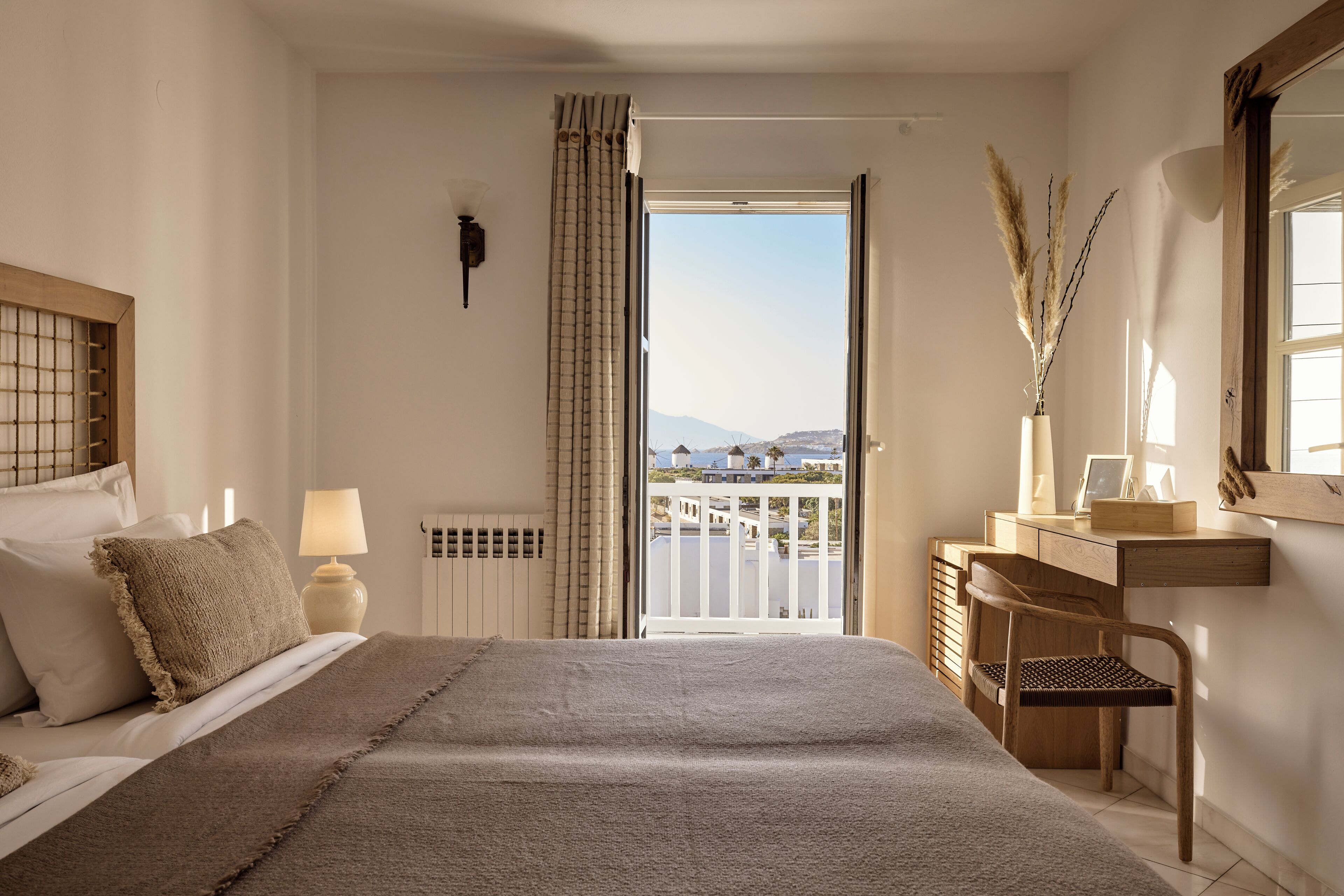 Quarto com cama, escrivaninha, cadeira, abajur, janela com cortinas e vista para edifícios.