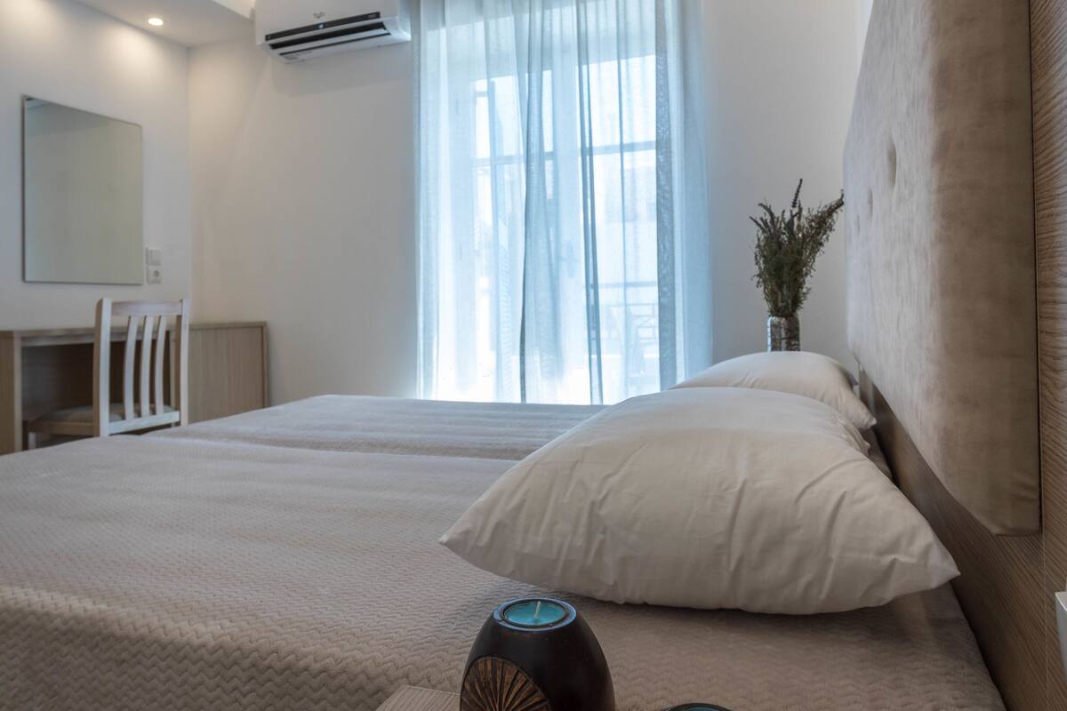Quarto de hotel moderno com duas camas, uma escrivaninha e uma janela grande com cortinas.