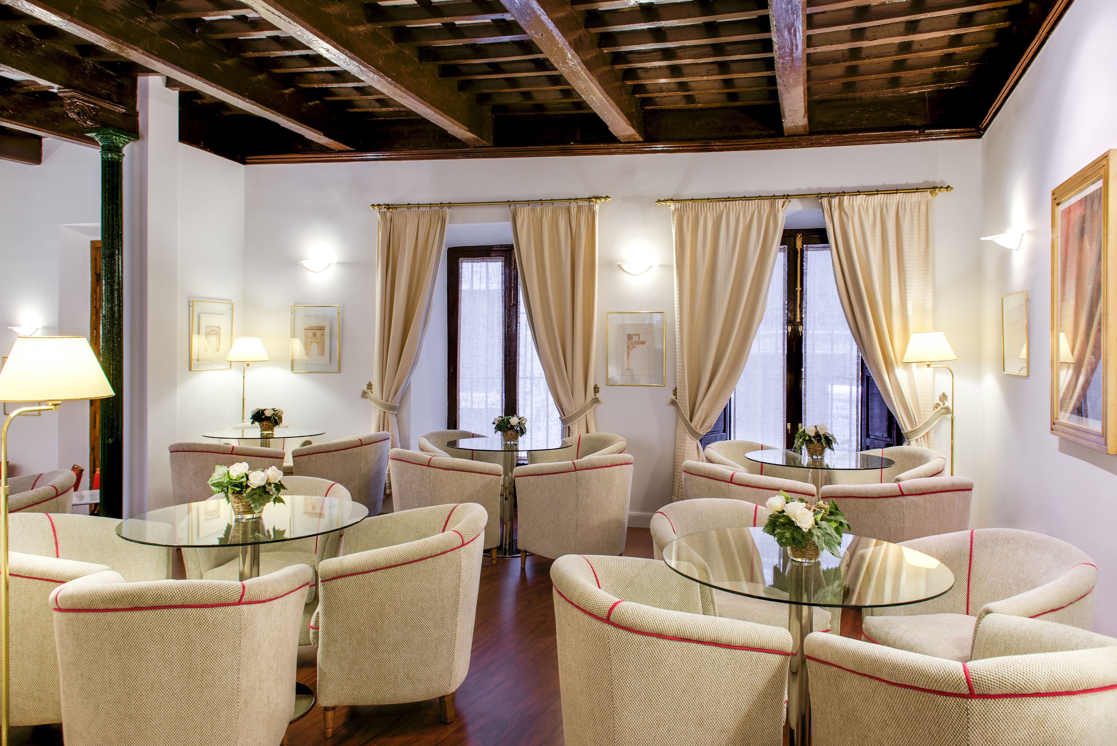 Um quarto com quatro mesas redondas de vidro, cada uma rodeada por quatro cadeiras bege estofadas com detalhes vermelhos. Há duas janelas com cortinas bege, e o quarto possui vigas de madeira no teto.