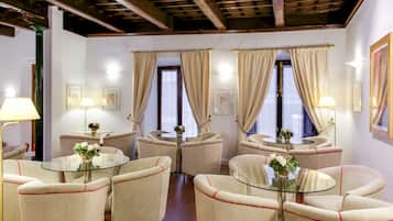 Um quarto com quatro mesas redondas de vidro, cada uma rodeada por quatro cadeiras bege estofadas com detalhes vermelhos. Há duas janelas com cortinas bege, e o quarto possui vigas de madeira no teto.