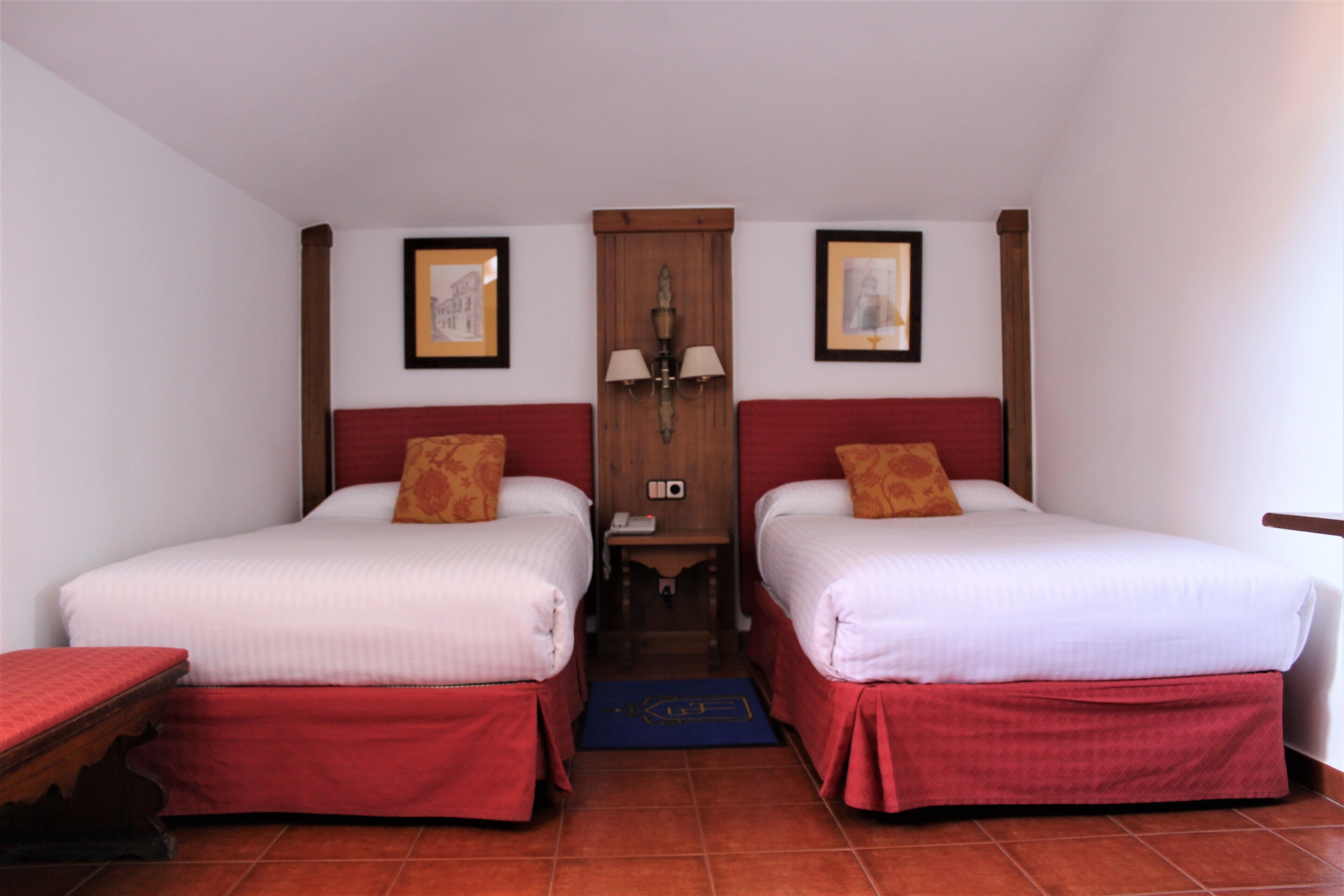 Quarto de hotel com duas camas, cada uma com roupa de cama vermelha e cobre correspondente. Há quadros nas paredes, uma porta de madeira e uma pequena mesa com abajur.