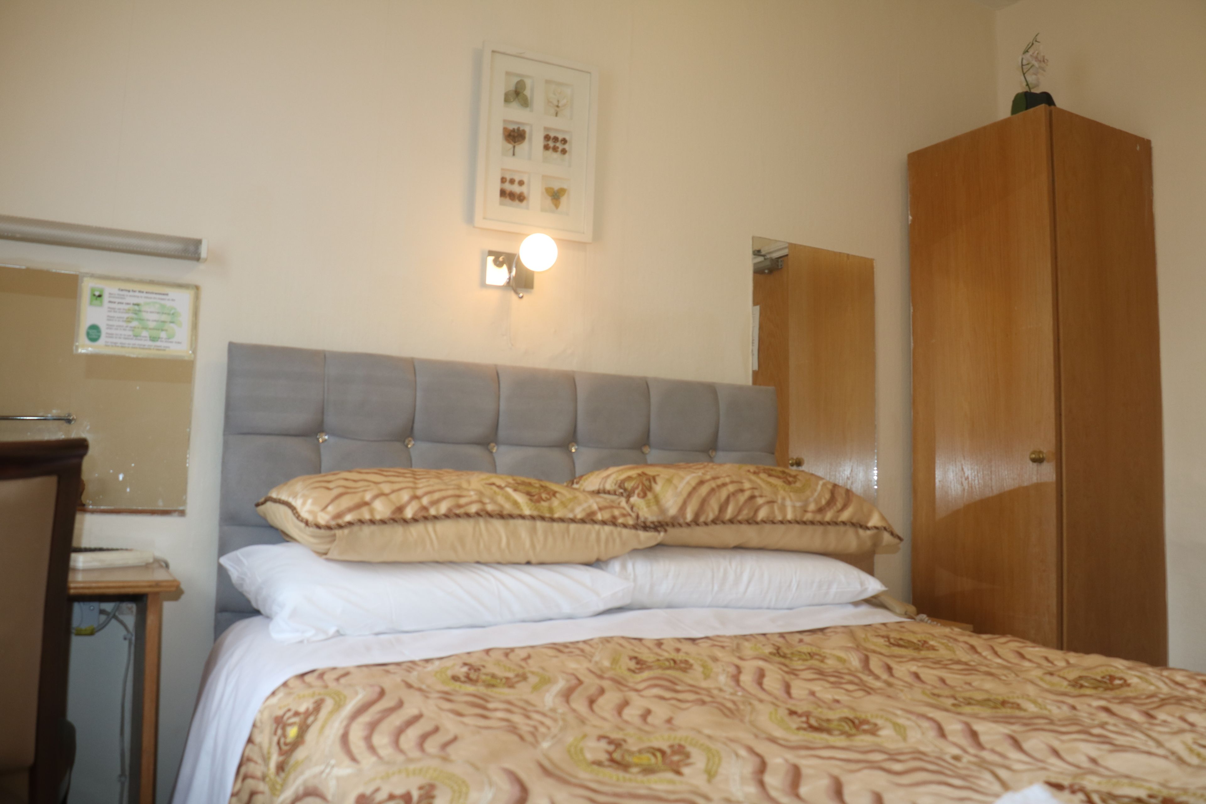 Um quarto com cama, mesa de cabeceira, luminária na parede, quadro emoldurado e guarda-roupa de madeira.