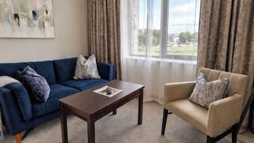 Sala de estar com sofá azul, poltrona bege, mesa de centro de madeira e vista pela janela com cortinas.