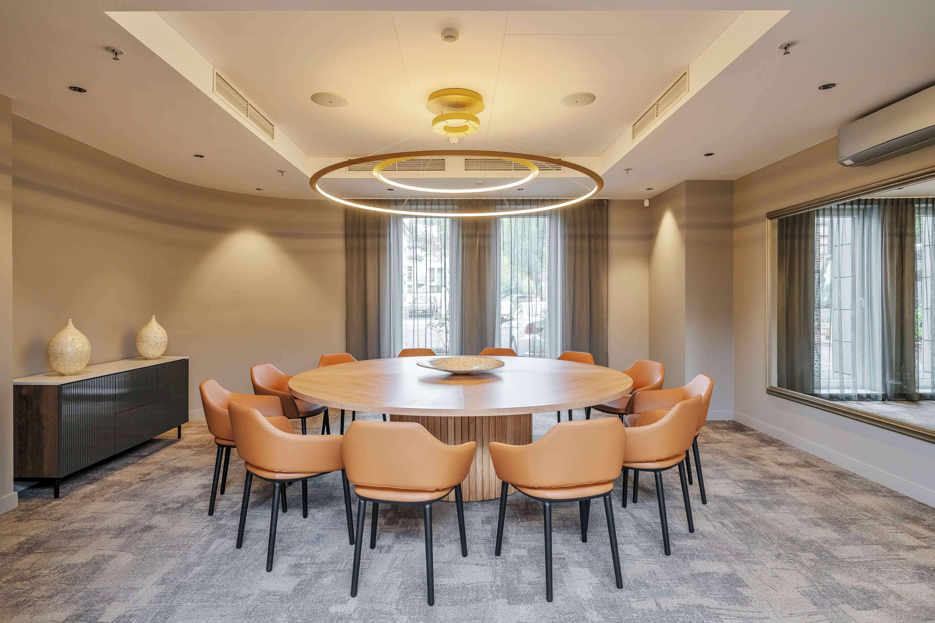 Uma sala de conferências com uma mesa redonda, oito cadeiras, um lustre central, um armário e janelas amplas.