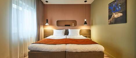 Uma cama de casal com cabeceira marrom, roupa de cama branca e um cobertor vermelho. Há duas luminárias de mesa, um espelho fixado na parede e um quadro de um veado na parede.