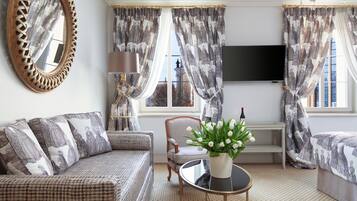 Uma sala de estar com sofá, poltrona e mesa redonda com um vaso de flores. Uma televisão está fixada na parede, e cortinas estampadas cobrem as janelas.