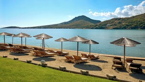 Uma praia com espreguiçadeiras e guarda-sóis de palha, um deck de madeira e vista para um lago e montanhas.