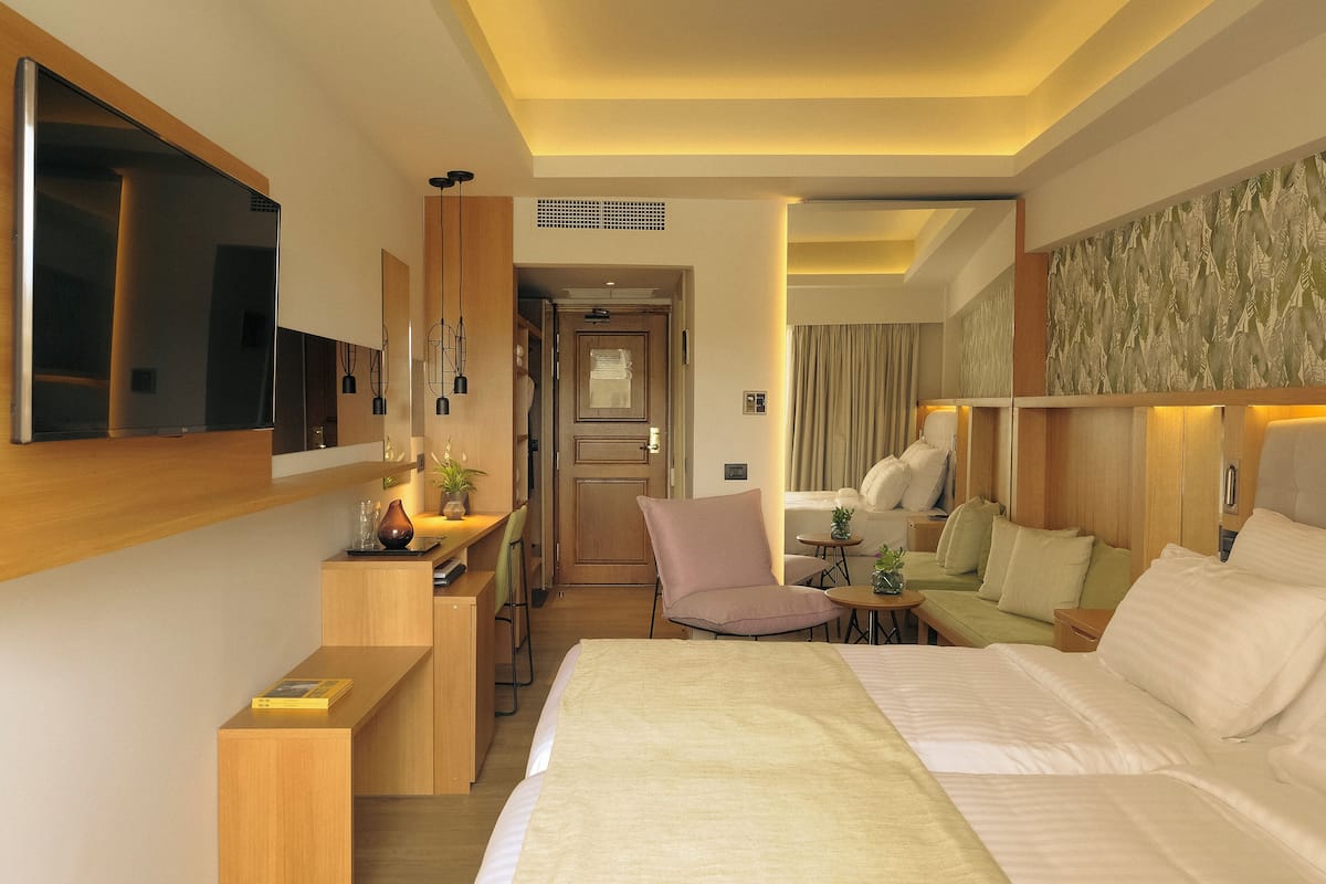 Quarto de hotel com uma cama grande, uma escrivaninha com cadeira, uma mesinha de cabeceira com abajur e uma televisão fixada na parede.