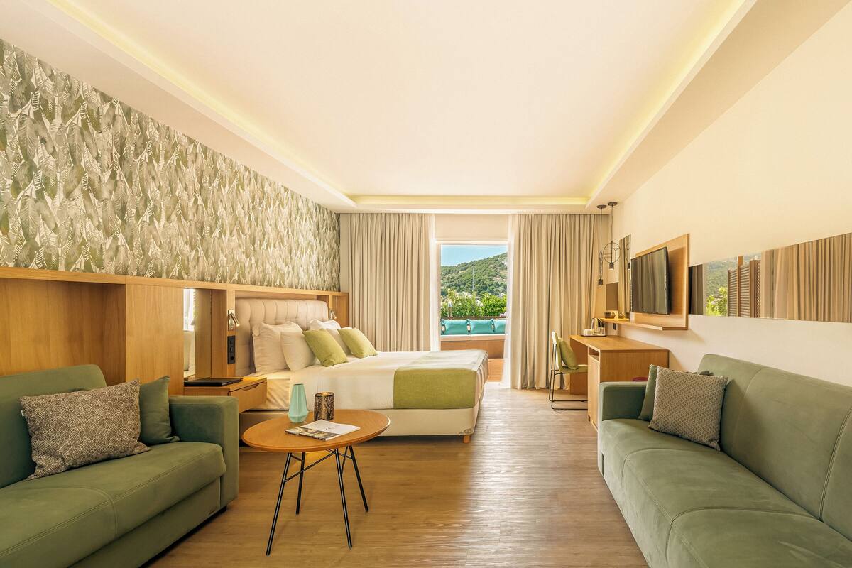 Quarto de hotel com uma cama, dois sofás verdes, uma mesa de centro de madeira, uma televisão e vista para uma paisagem verde.